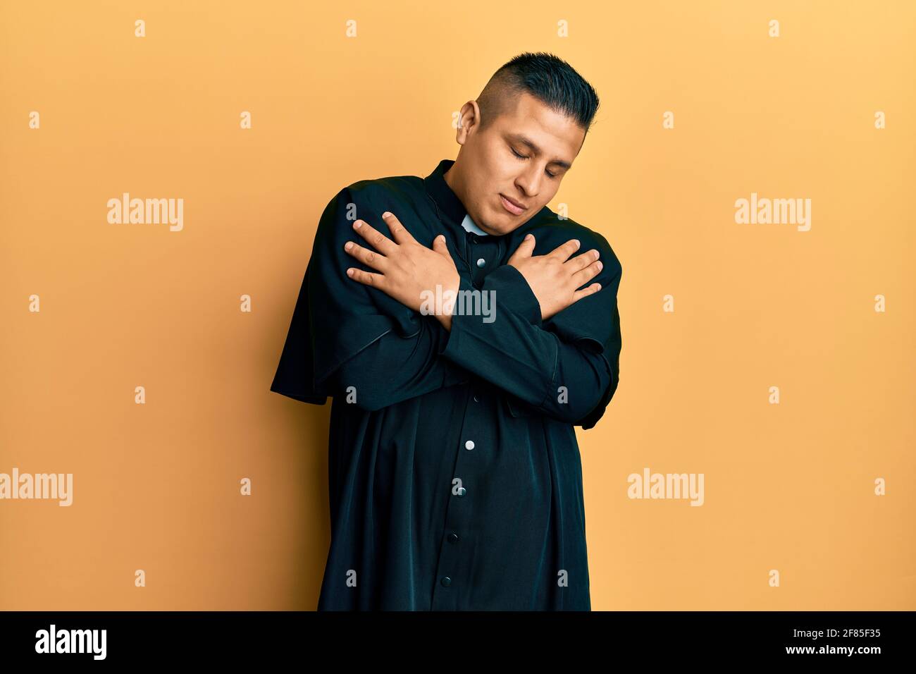 Young latin priest man standing over yellow background hugging oneself ...