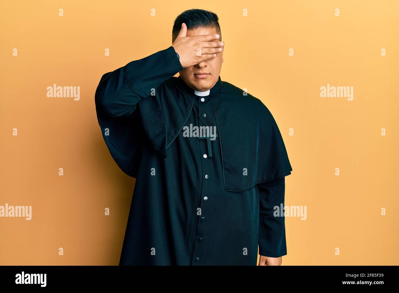Young latin priest man standing over yellow background covering eyes ...