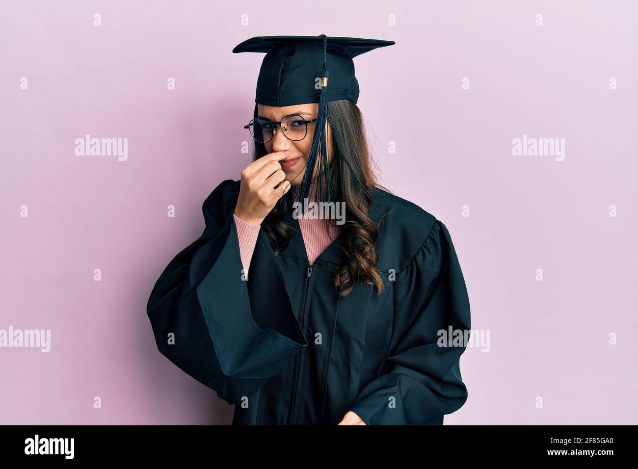 Young hispanic woman wearing graduation cap and ceremony robe smelling ...