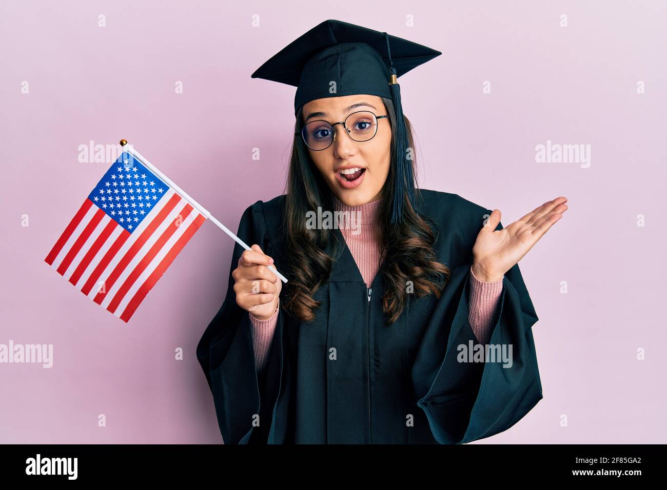 Young hispanic woman wearing graduation cap and ceremony robe holding ...