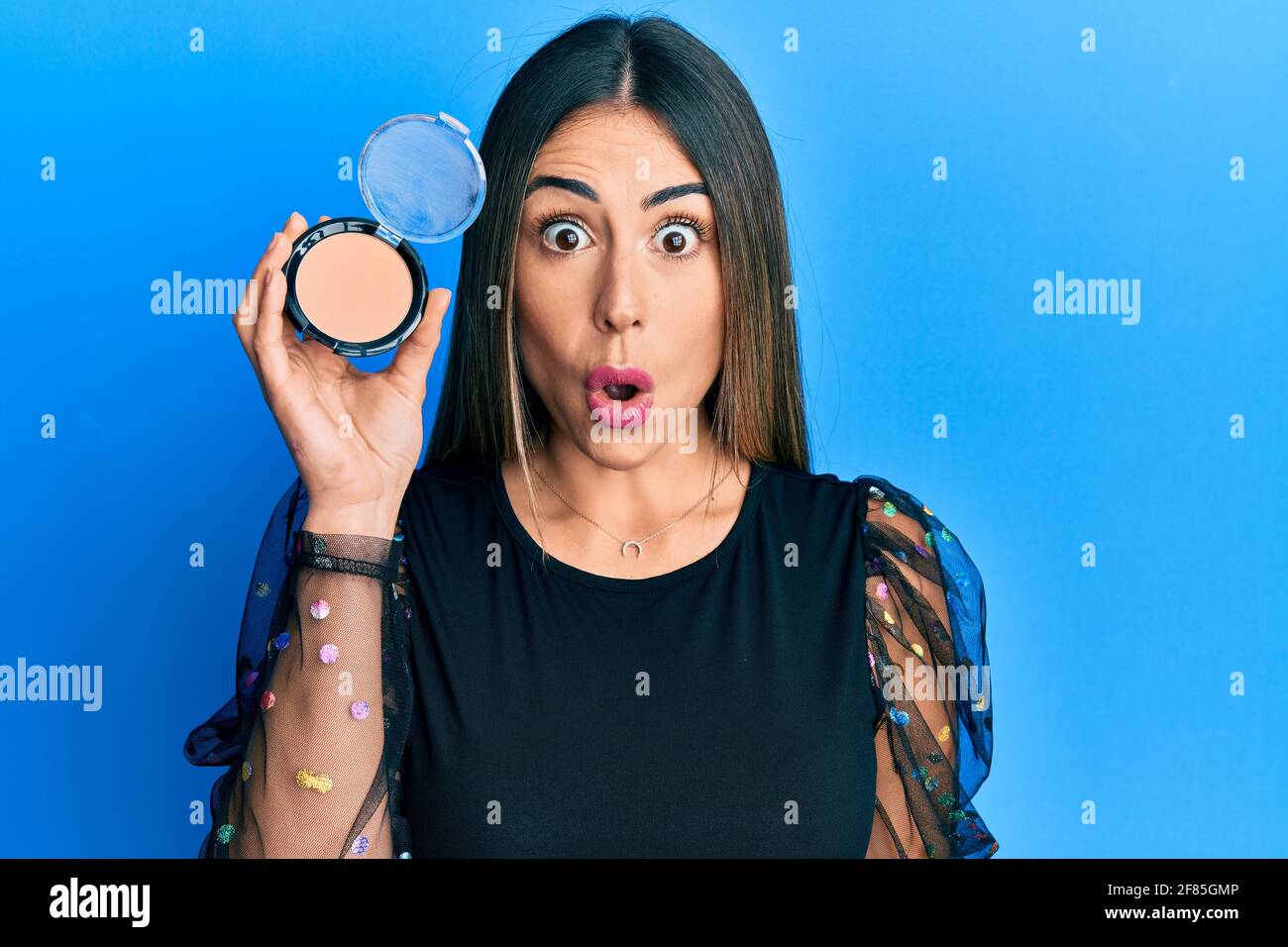 Young hispanic woman holding makeup powder scared and amazed with open ...