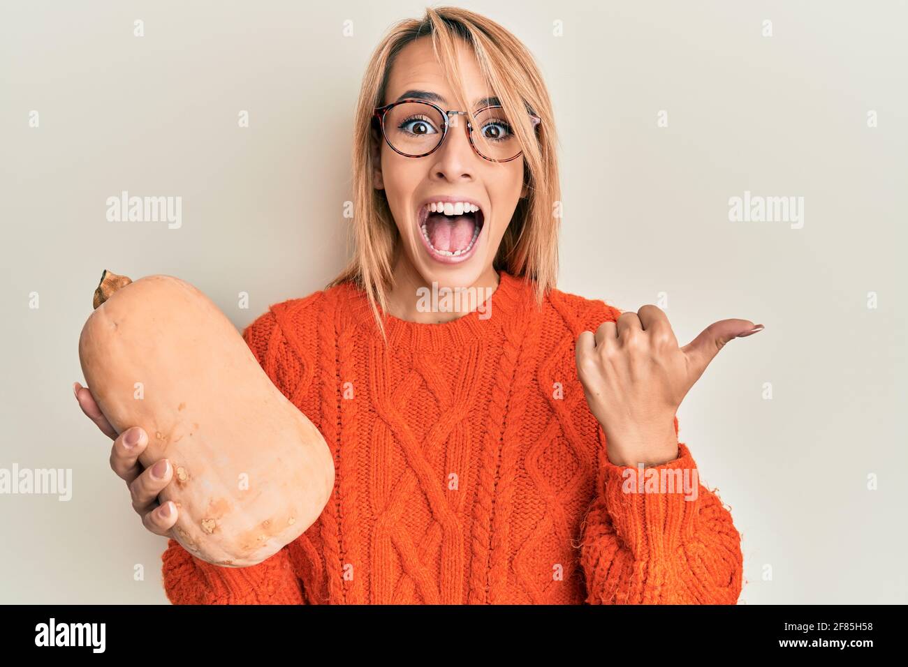Beautiful blonde woman holding healthy fresh pumpkin pointing thumb up ...