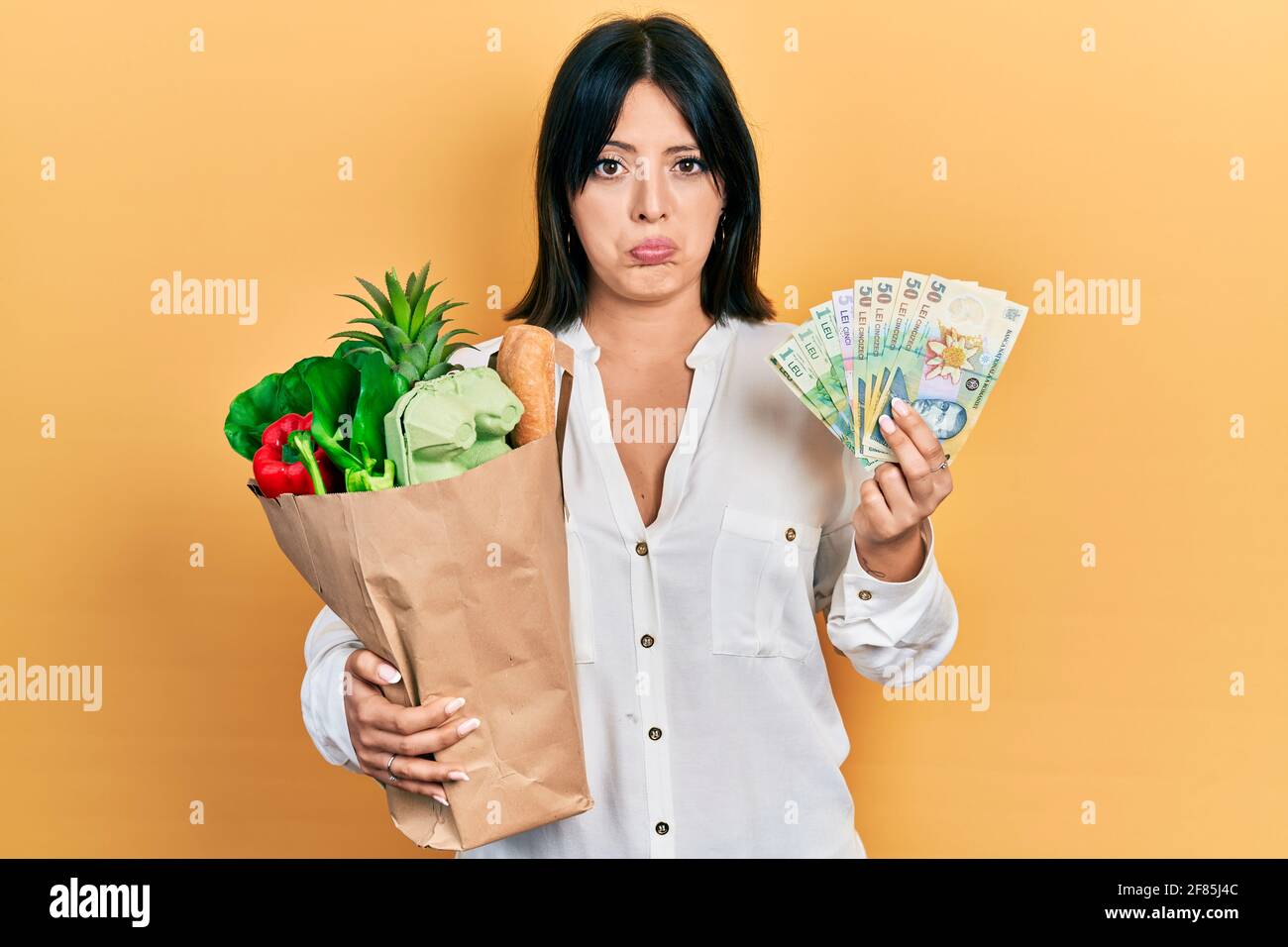 Young hispanic woman holding groceries and romania leu banknotes ...