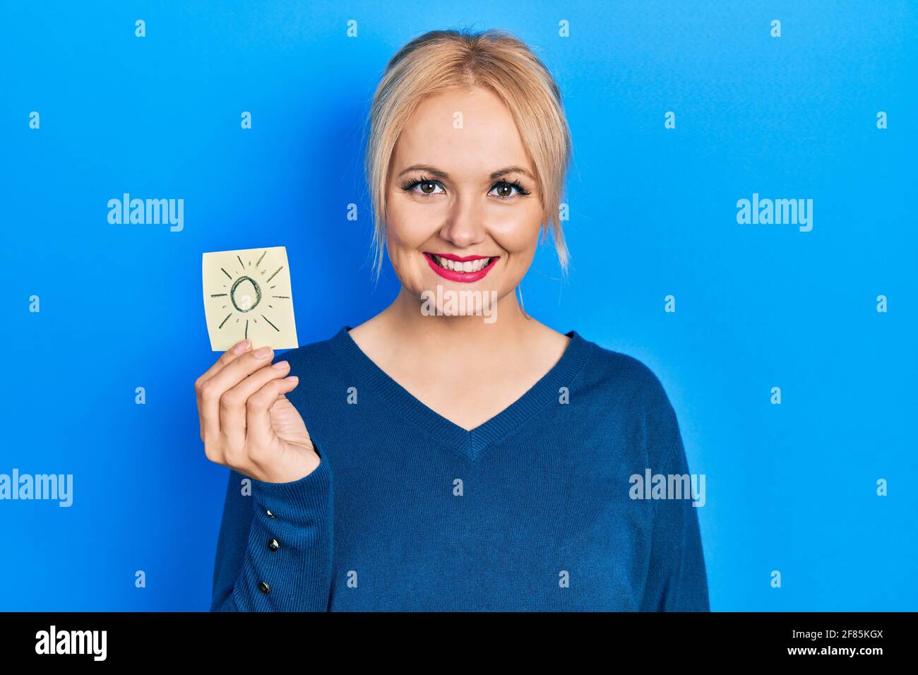 Young blonde woman holding sun draw reminder looking positive and happy ...