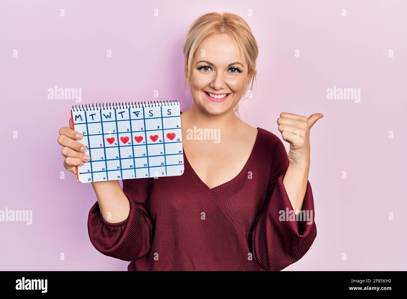 Young blonde woman holding heart calendar pointing thumb up to the side ...