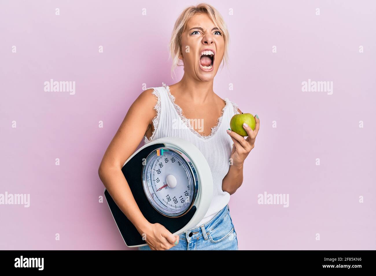 Young blonde girl holding weight machine and green apple angry and mad ...