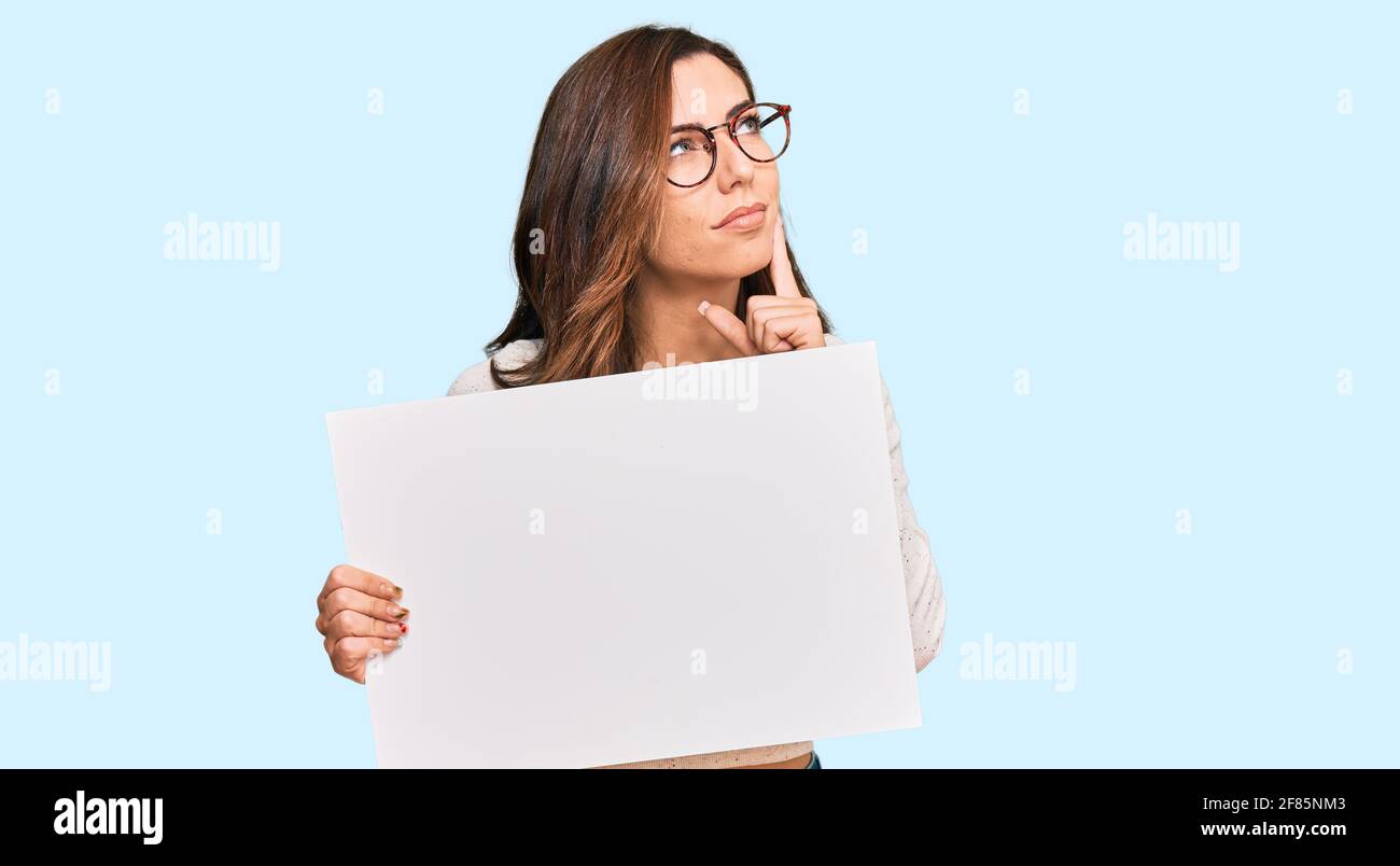 Young brunette woman holding blank empty banner serious face thinking ...