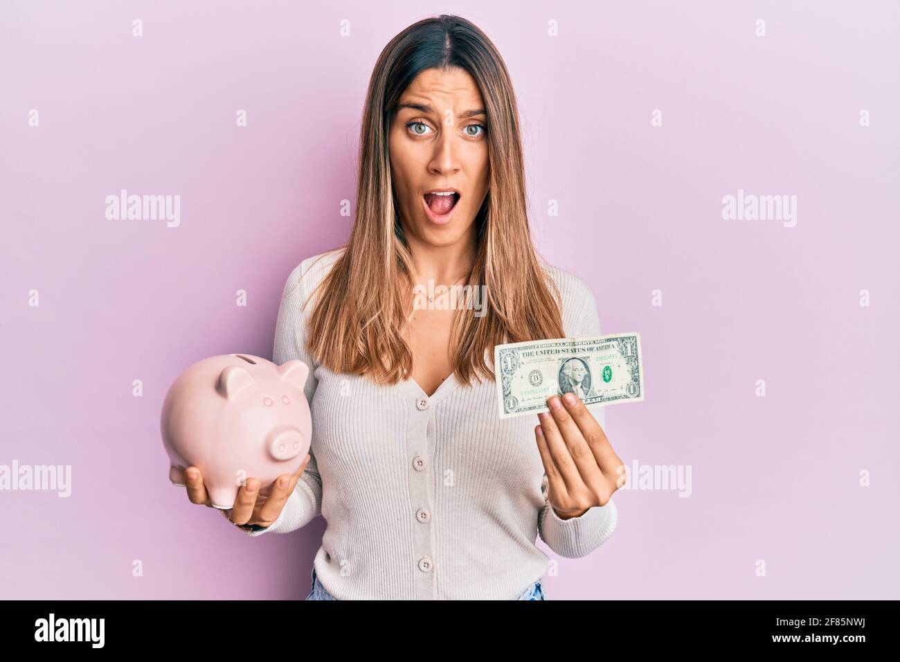 Brunette young woman holding one dollar banknote and piggy bank afraid ...
