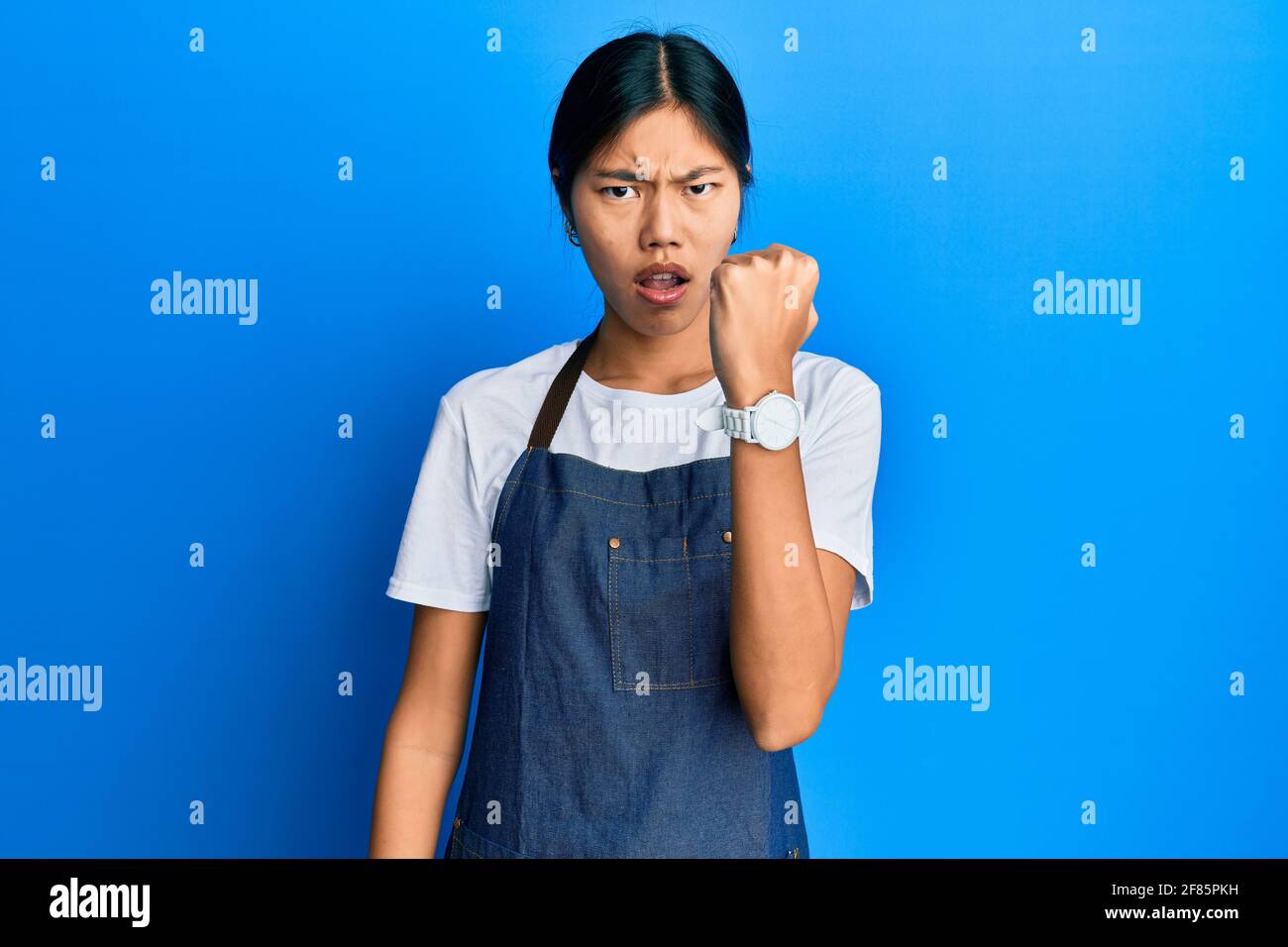 Young chinese woman wearing waiter apron angry and mad raising fist ...