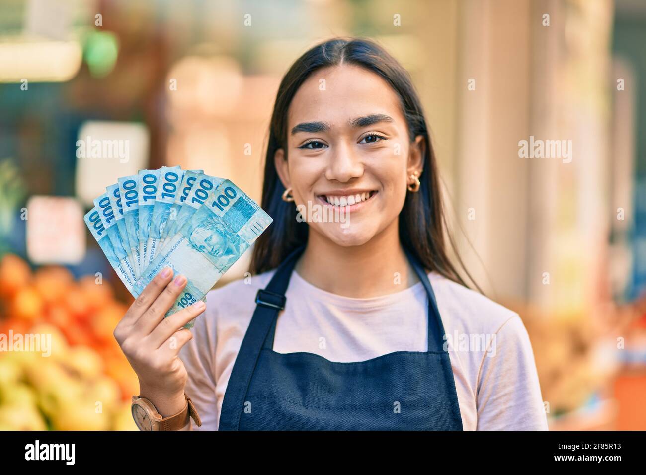 Young latin shopkeeper girl smiling happy holding brazil real banknotes ...