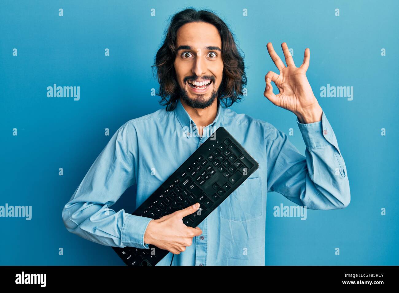 Young hispanic man holding keyboard doing ok sign with fingers, smiling ...