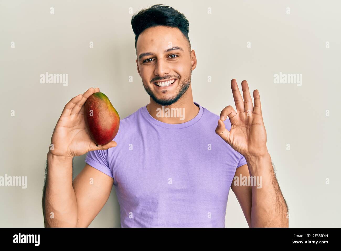 Young arab man holding mango doing ok sign with fingers, smiling ...