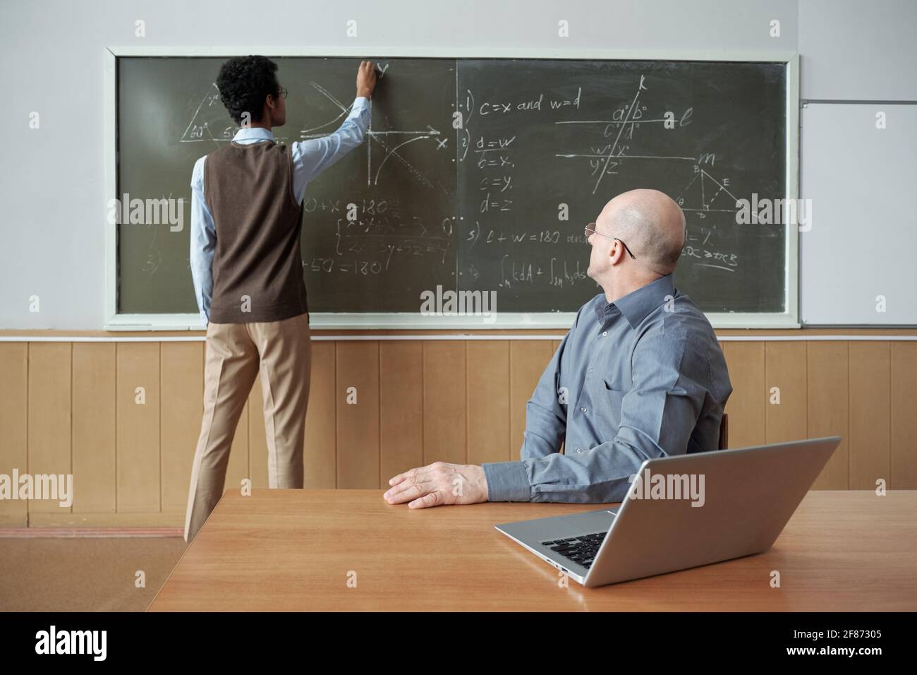 Mature bald professor sitting by desk and looking at student standing ...