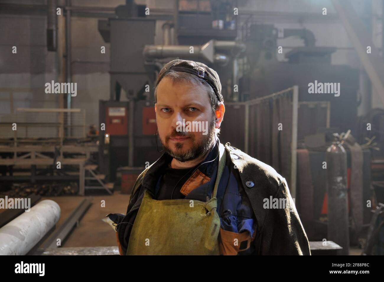 A worker with a beard in overalls in a production room at a factory ...