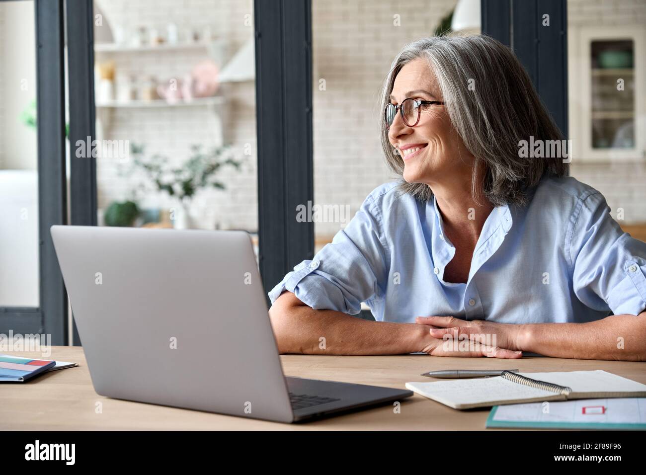 Middle aged woman sitting at home with computer smiling dreamingly ...