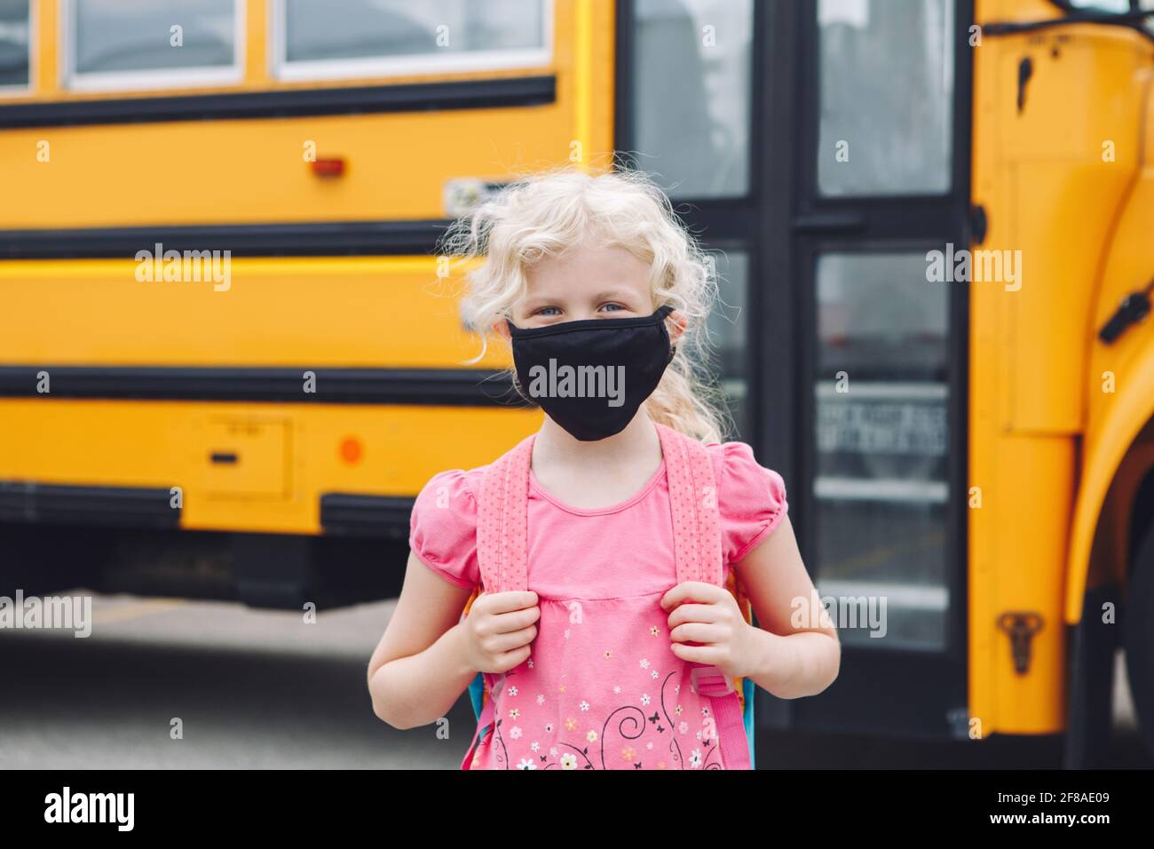 Happy girl student in cloth face mask by yellow school bus outdoors ...