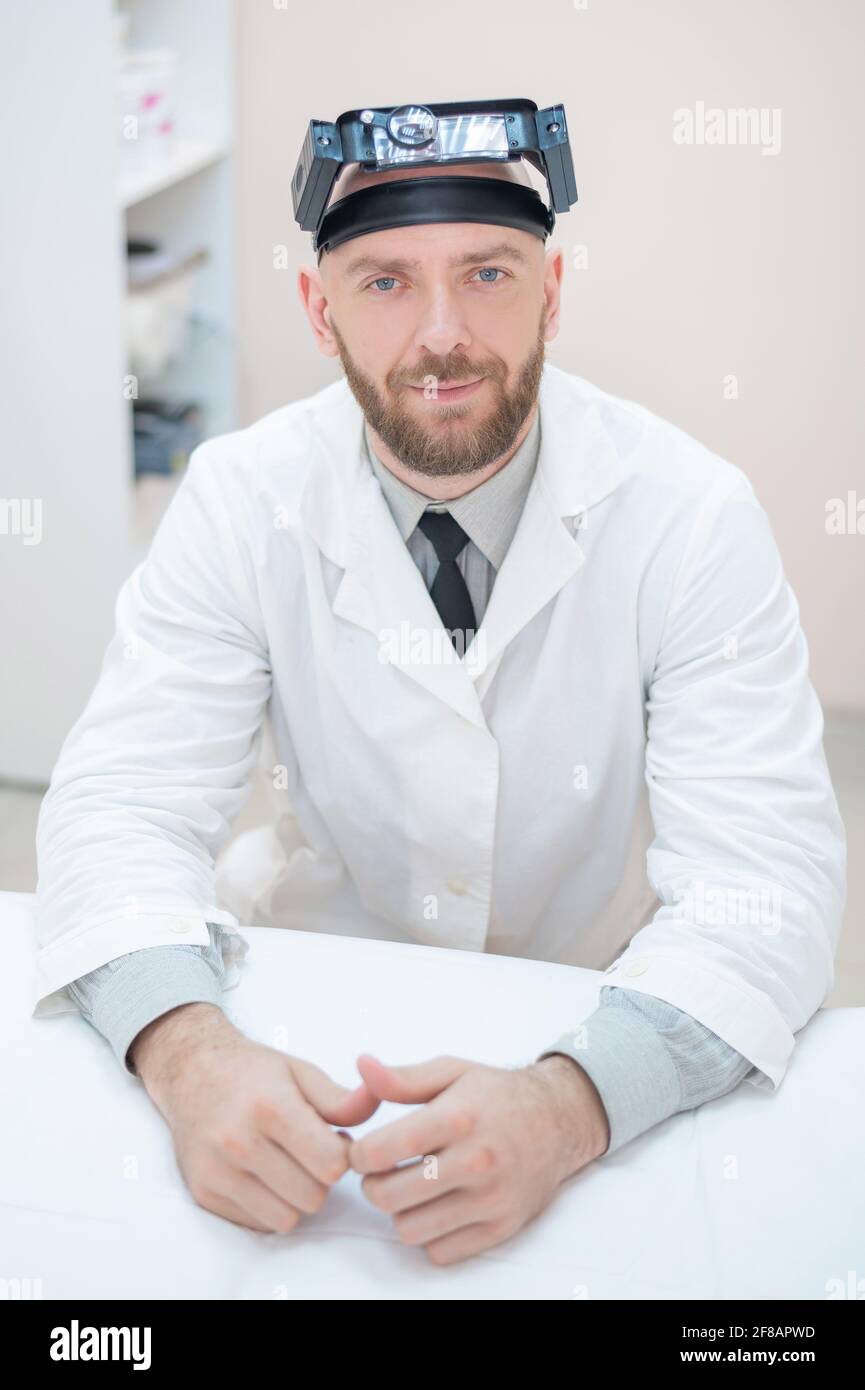 Male bearded doctor in a medical coat and with a magnifying head strap ...