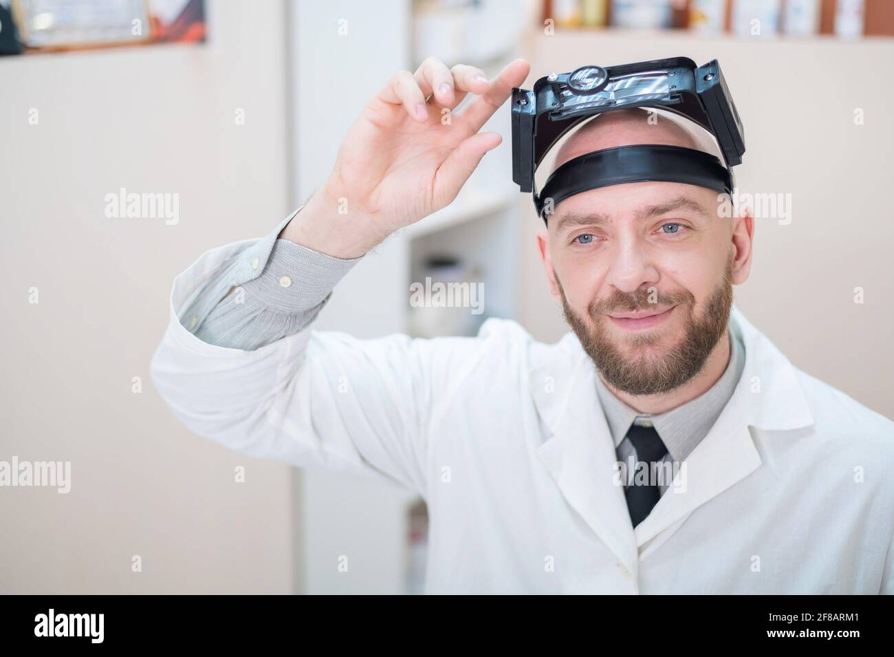 Male bearded doctor in a medical coat and with a magnifying head strap ...