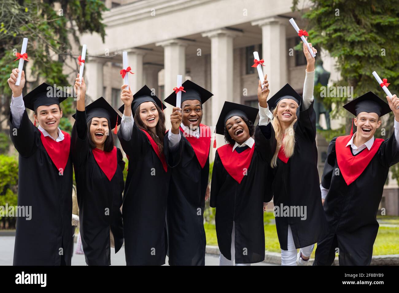 Cheerful multiracial students in graduation costumes raising diplomas ...