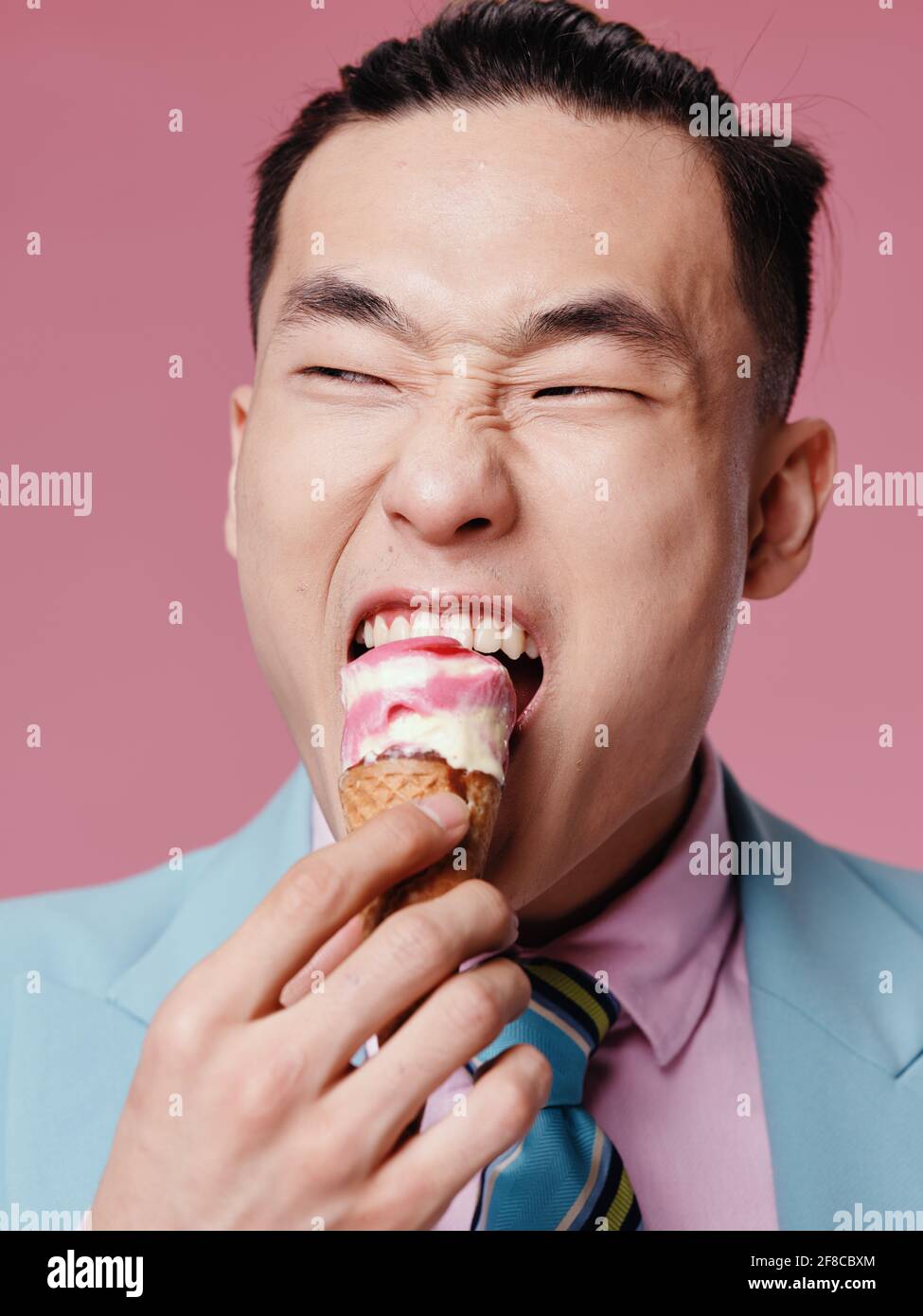 A man squinting his eyes eats ice cream in a suit on a pink background ...