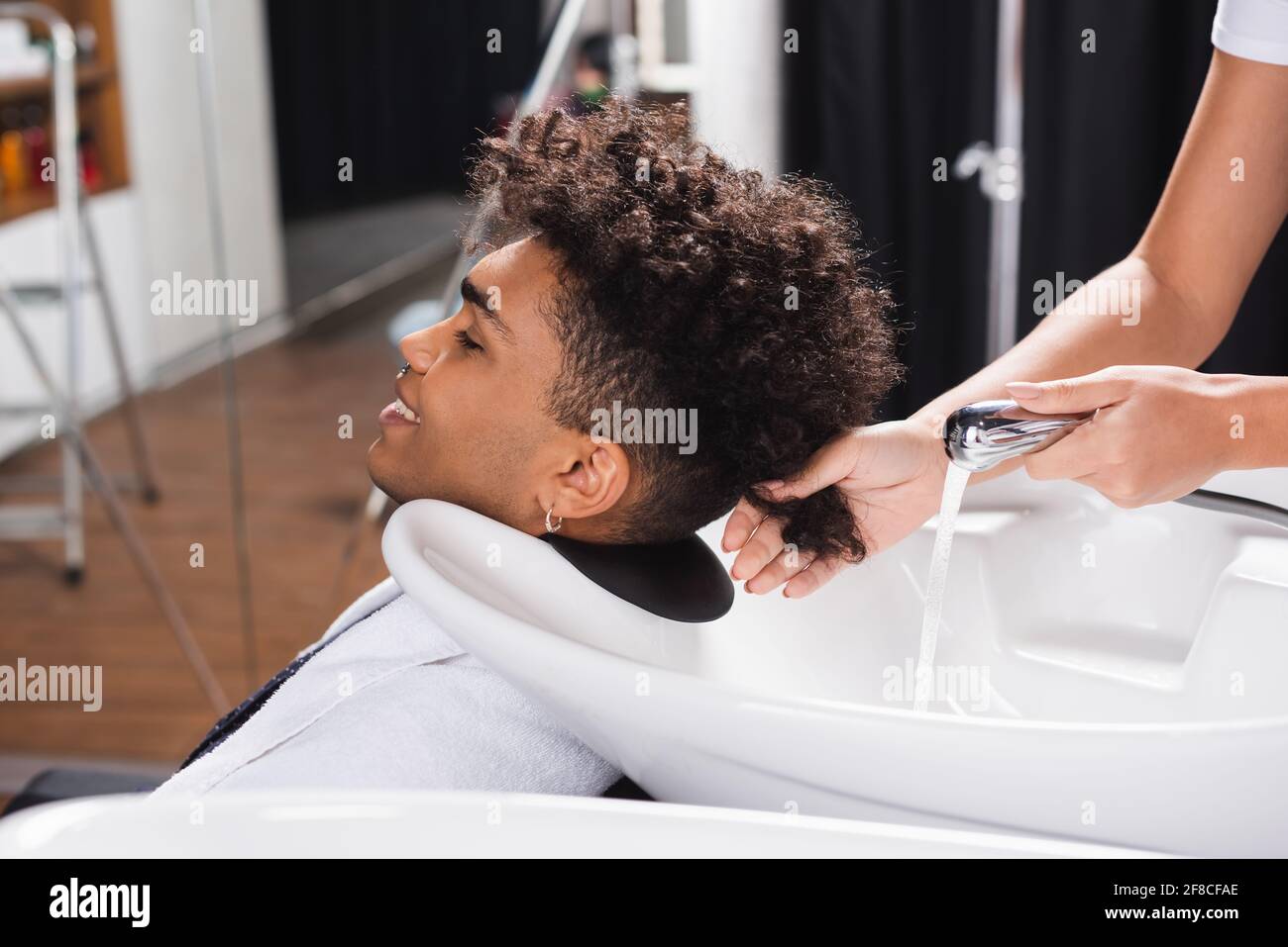 Smiling african american client sitting near hairdresser with shower ...
