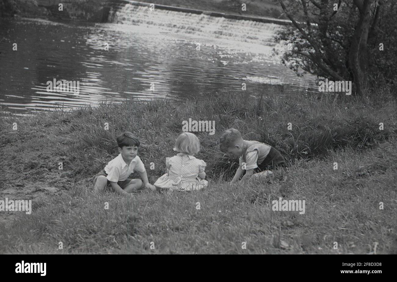 1960s, historical, three young children together on a grassy riverbank ...