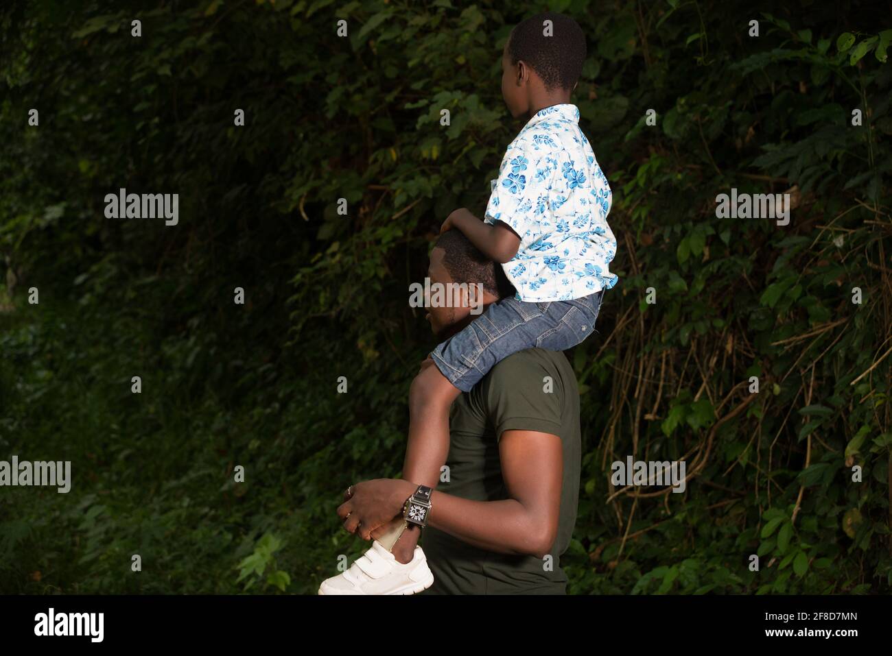 a young man standing in a park carrying his child on his neck, looking ...