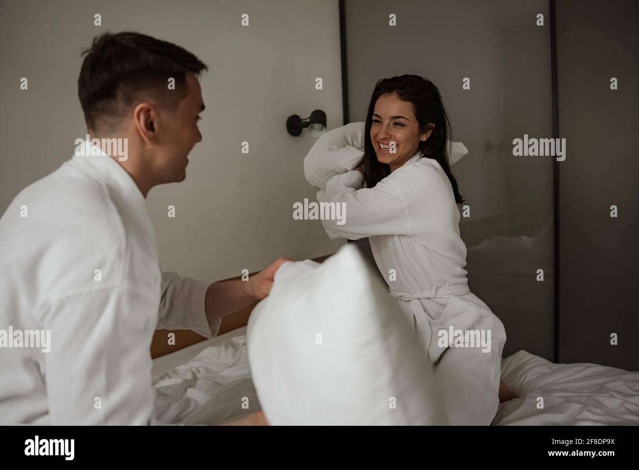Morning happy young couple in white bathrobes are beating pillows in ...