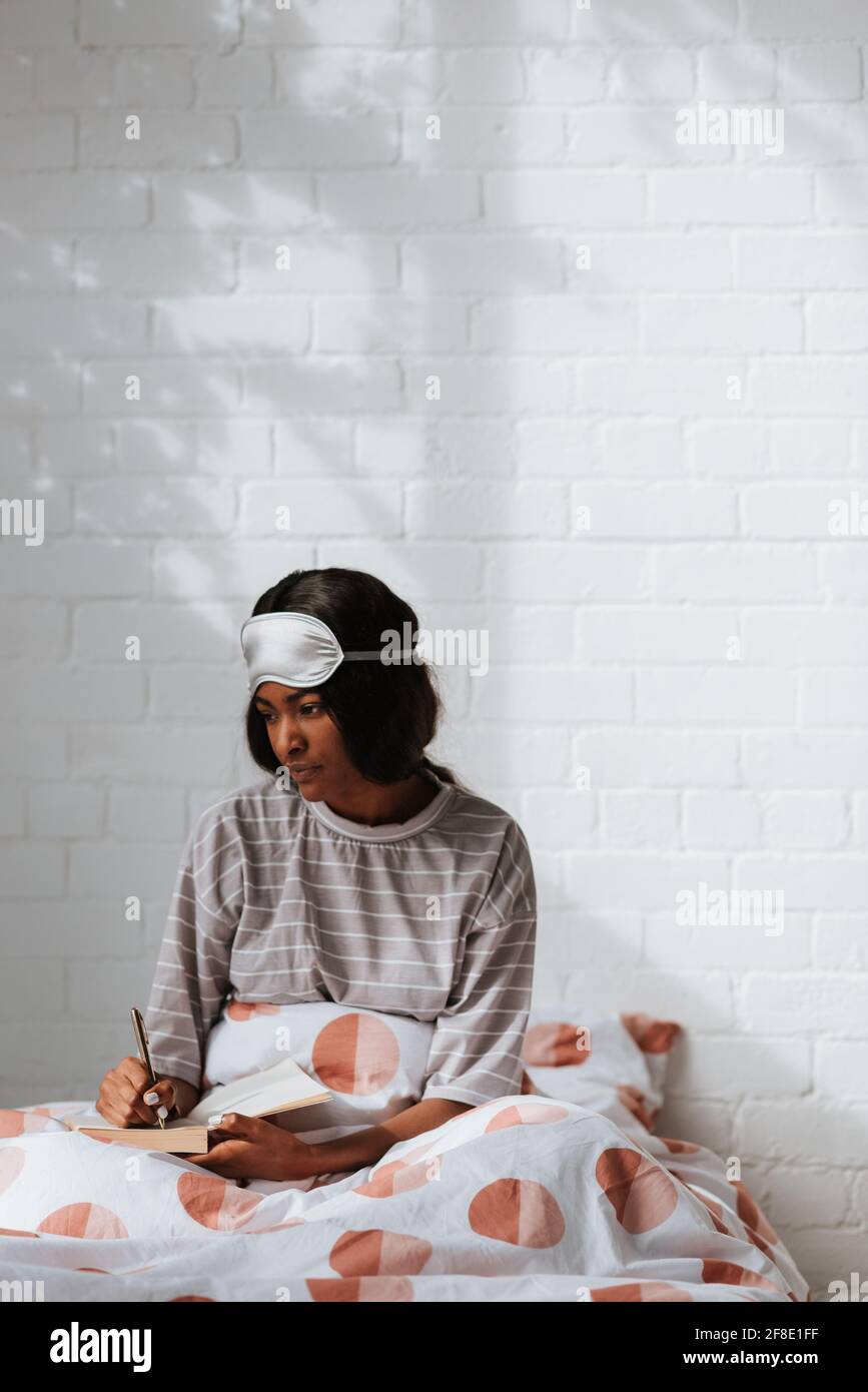 Thoughtful young African female writing notes while sitting on the bed ...
