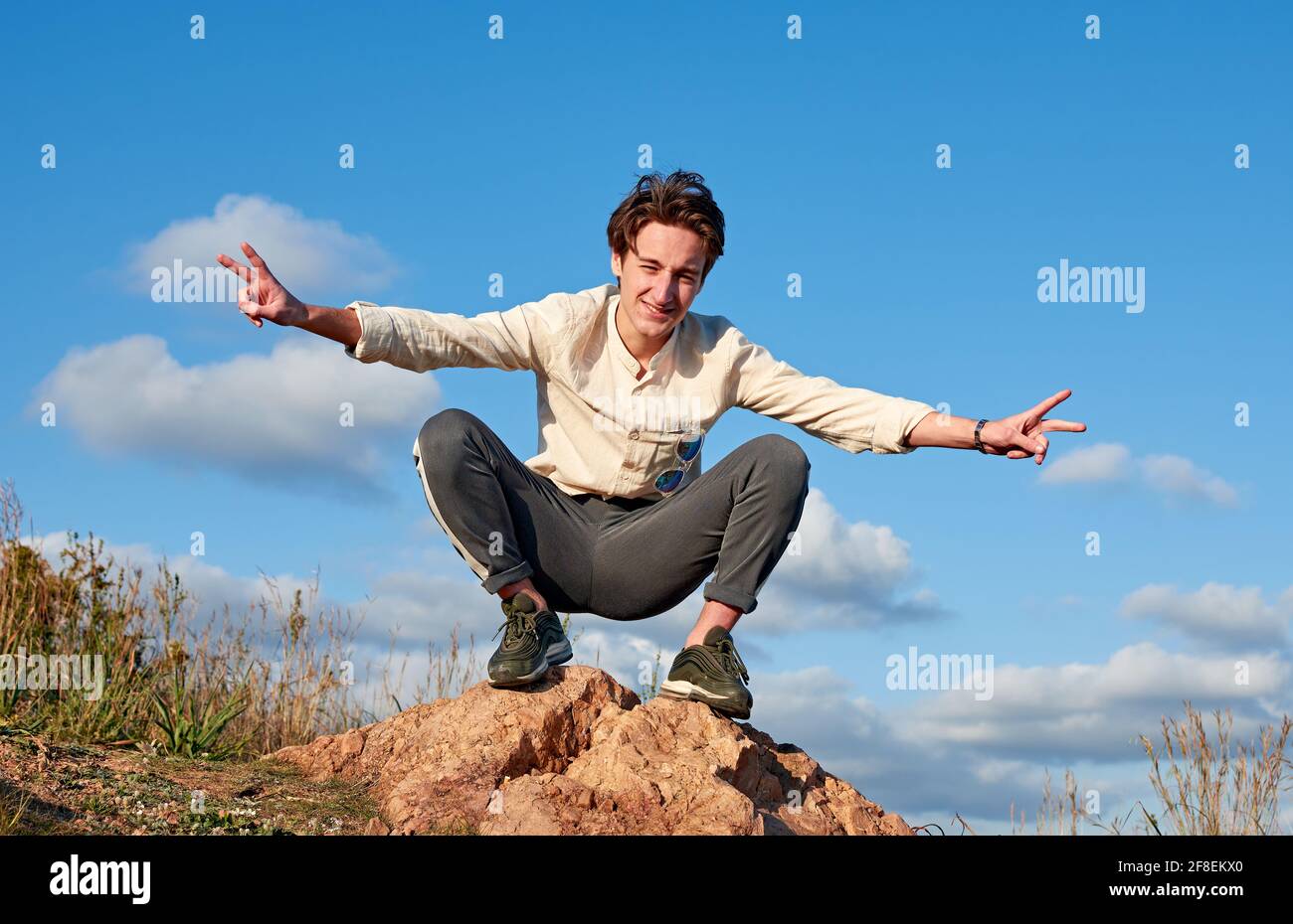 Cheerful young Spanish male squatting on a rock showing the peace sign ...