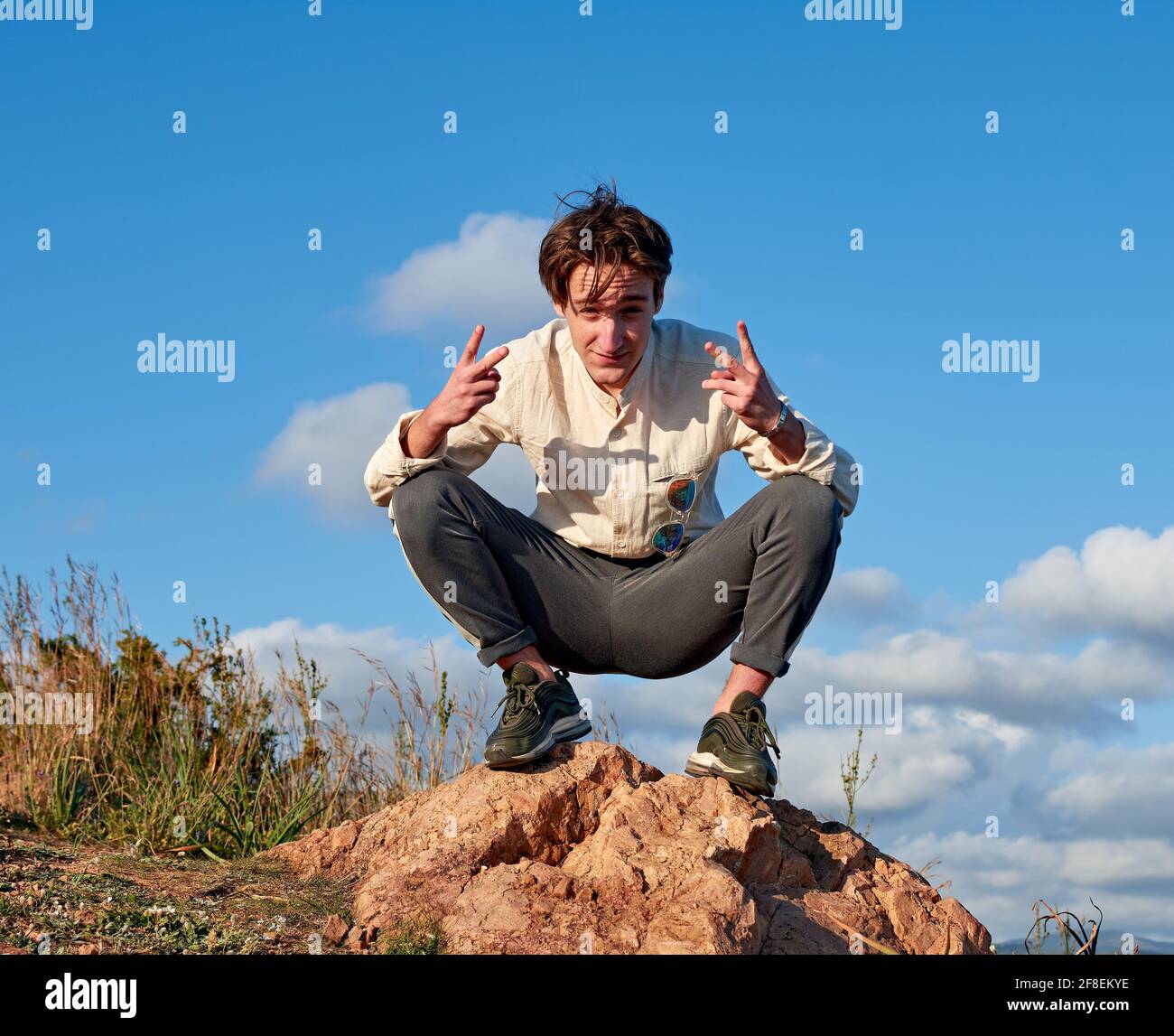 Stylish young Spanish male squatting on a rock showing the peace sign ...