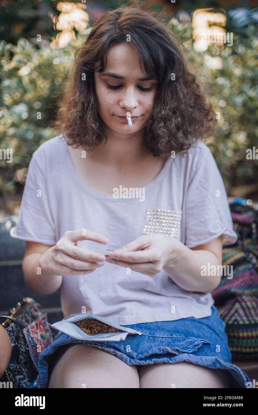 Addicted young woman sitting on park bench and rolling cigarette ...
