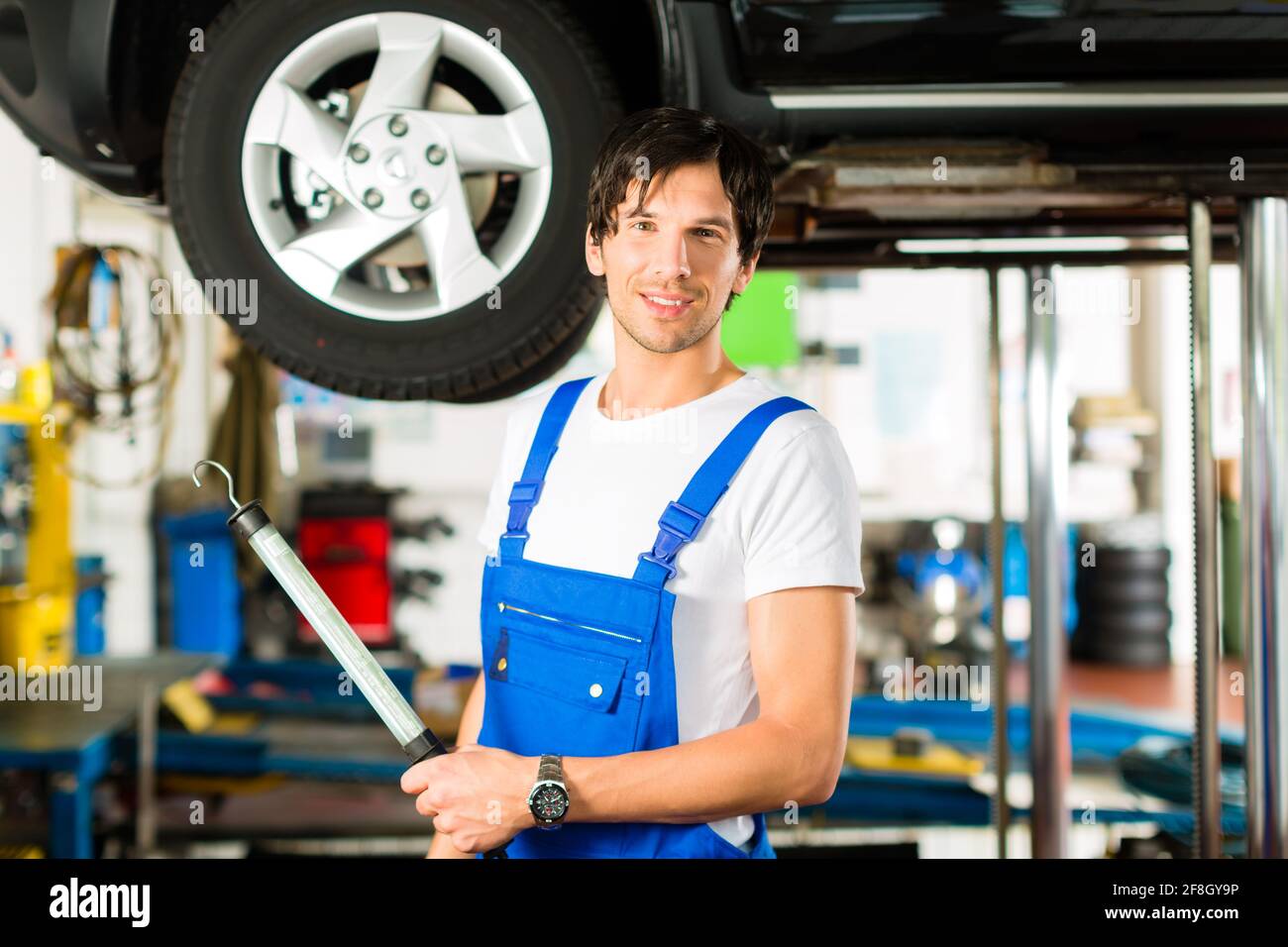 Young man in blue overall - mechanic - working with lamp on jacked car ...
