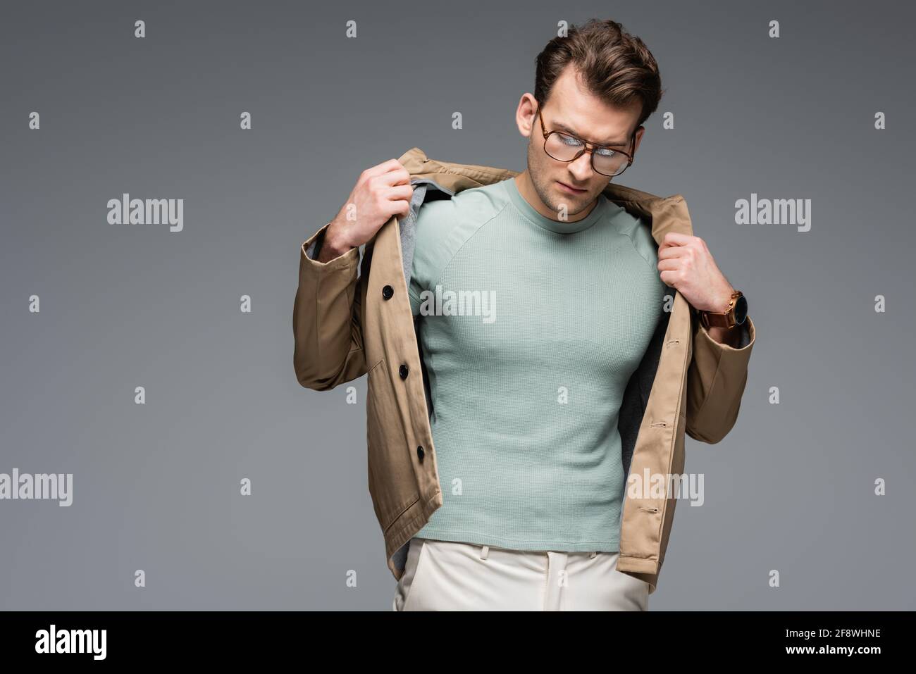 trendy man in glasses adjusting jacket while posing isolated on grey ...