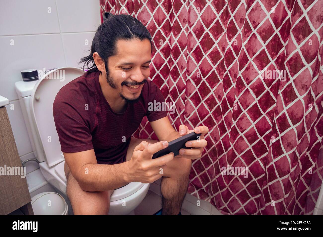 Young latin man smiling on the toilet using a smart phone at the ...