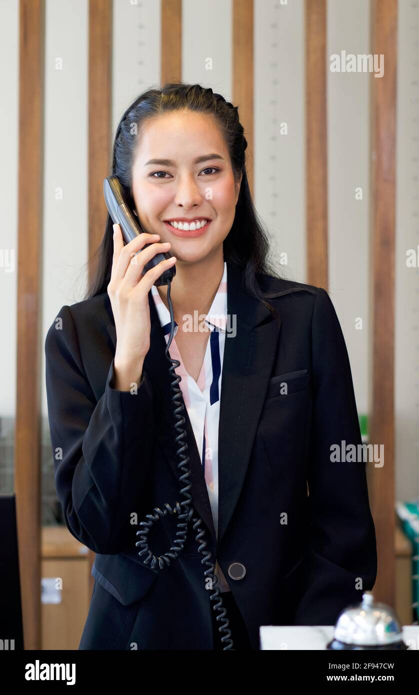 Asian hotel receptionist in black suit answer the phone while smiling ...