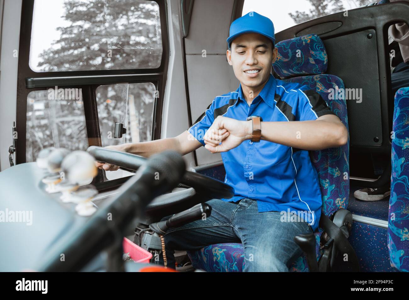 A male bus driver in blue uniform looks at his watch while sitting ...