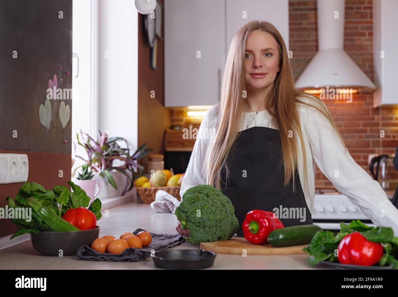 A young blonde happy lady standing in the kitchen while cooking healthy ...