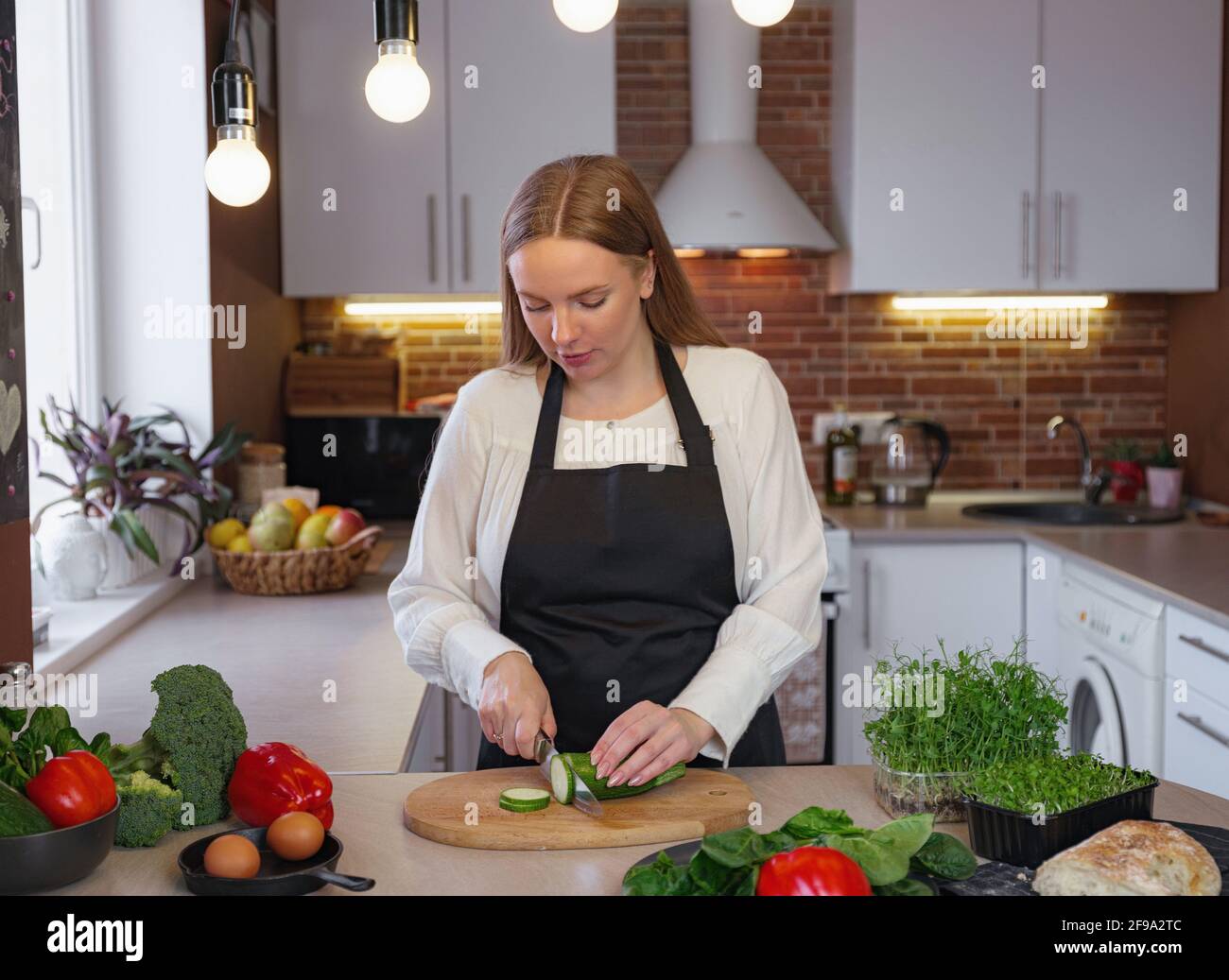 A young blonde happy lady standing in the kitchen while cooking healthy ...