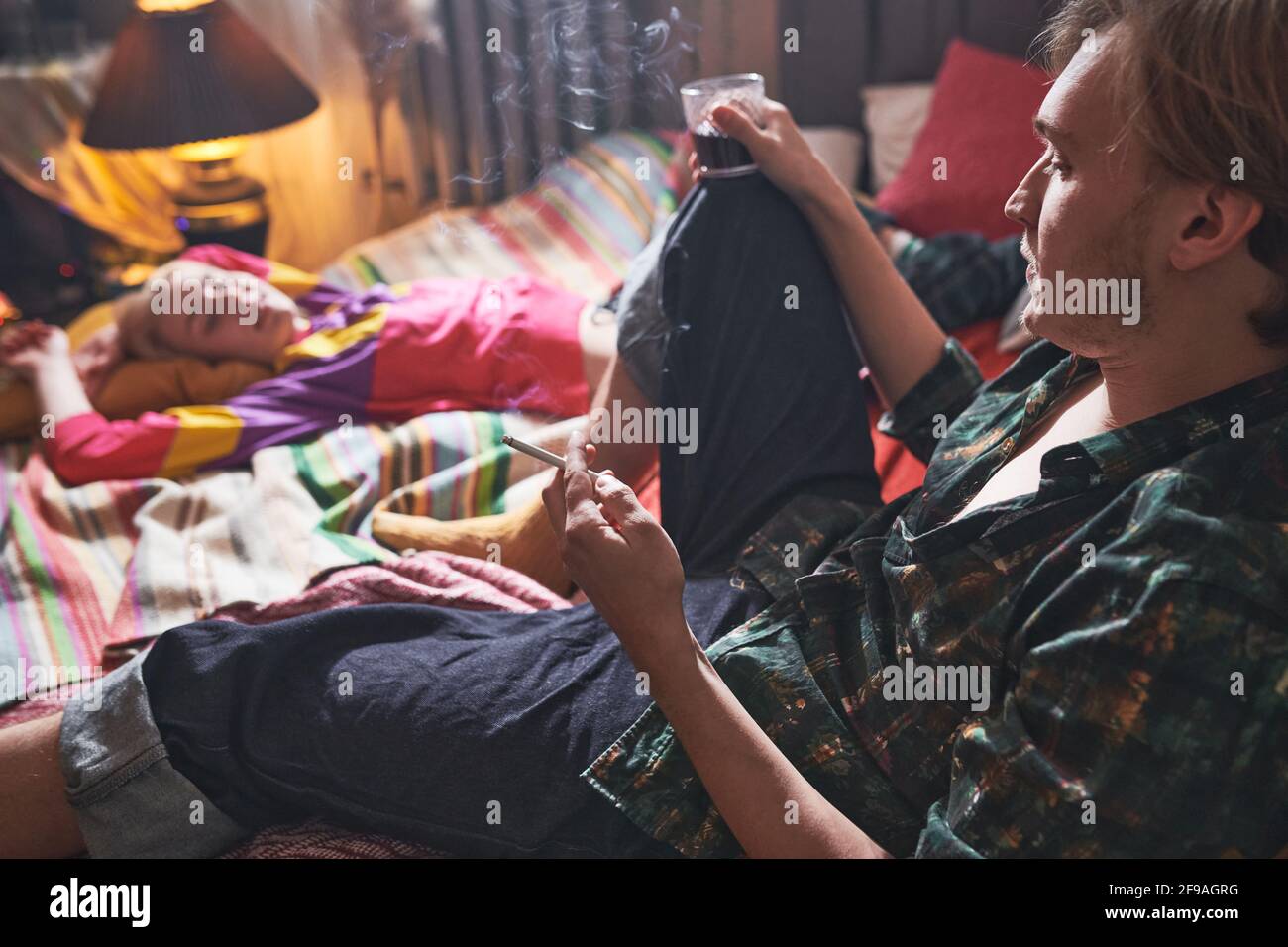 Young man smoking a cigarette and drinking wine sitting on the bed with ...