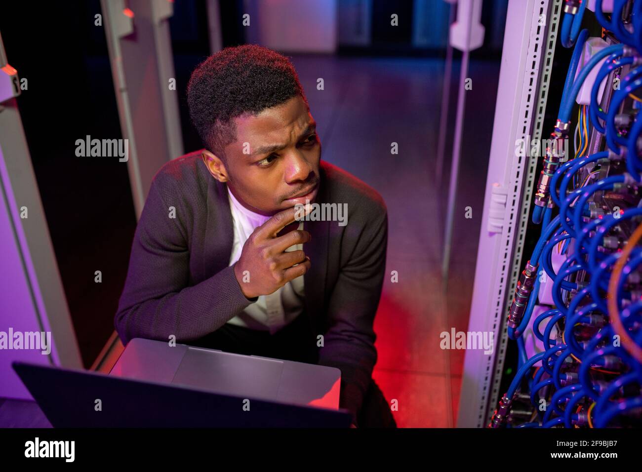 Thoughtful young black server engineer looking at cables in server ...