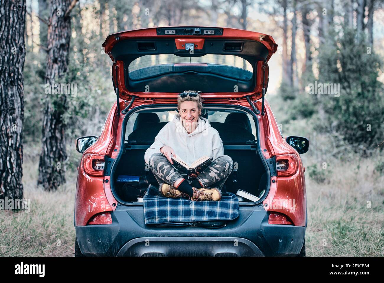 Young adult caucasian woman reading a book on the open trunk of a car ...