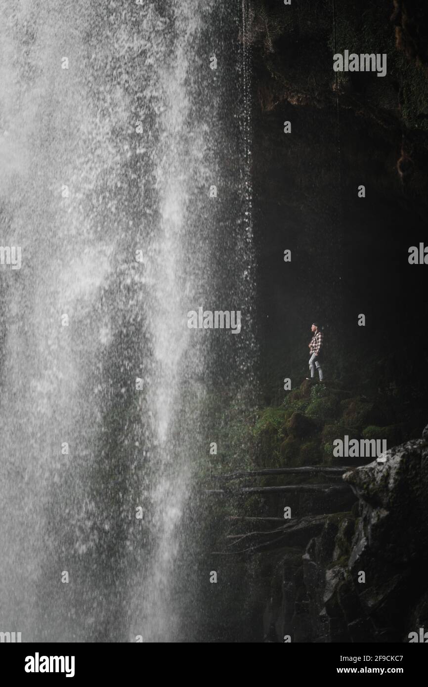 Vertical shot of an adult Caucasian man standing near a waterfall in a ...