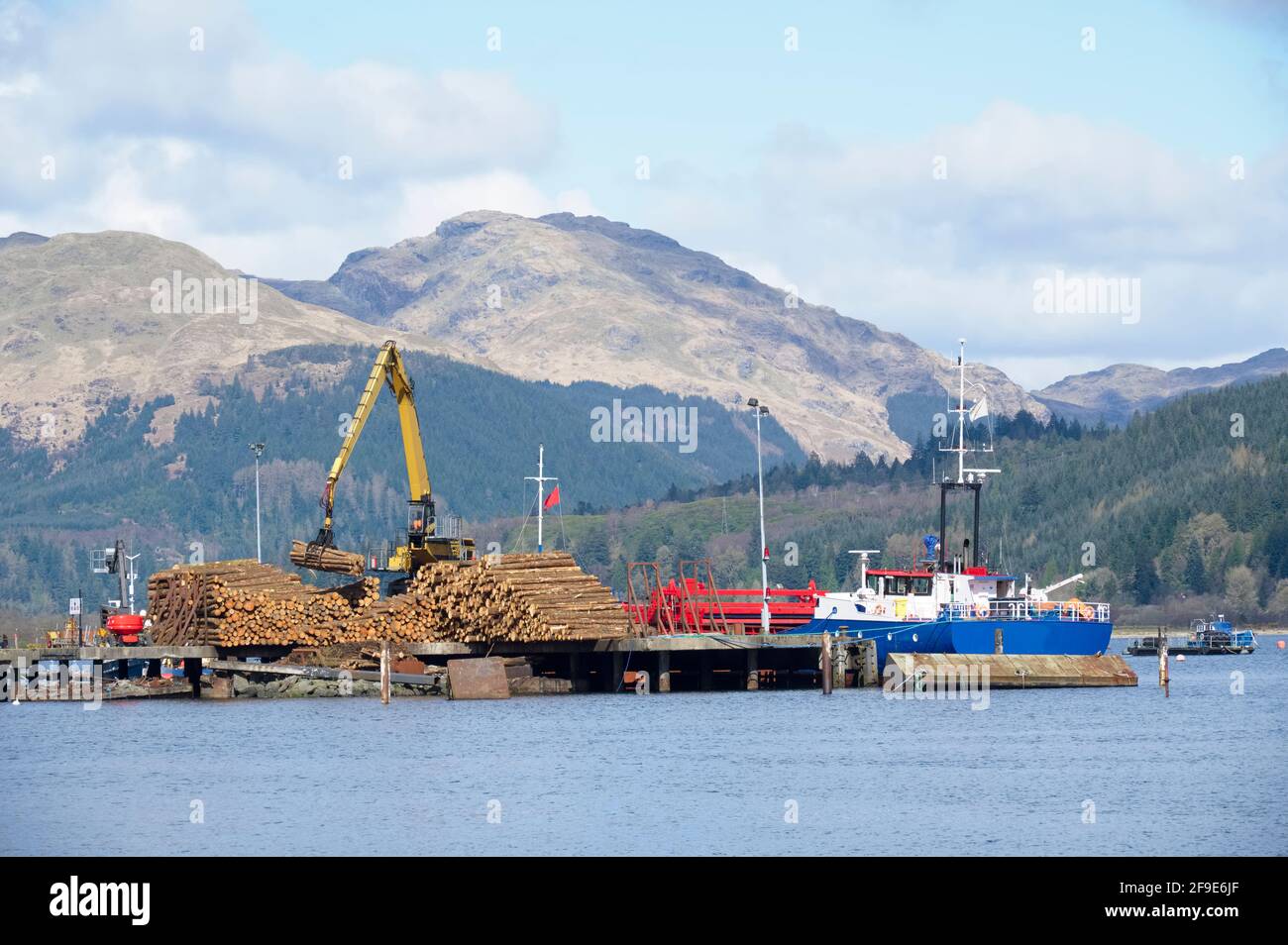Crane lifting wood logs from stack to ship in sea loch for ...