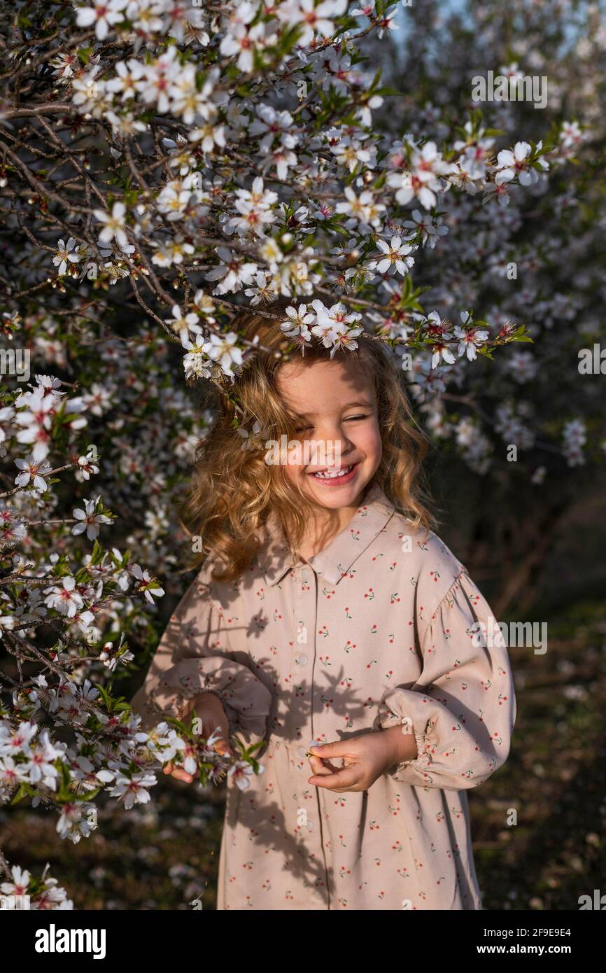 Adorable smiling little child in dress standing near blossoming tree ...