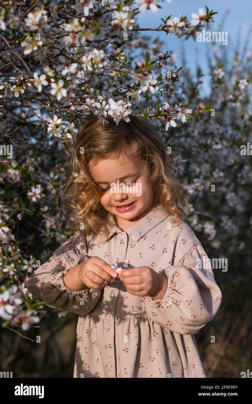 Adorable smiling little child in dress standing near blossoming tree ...