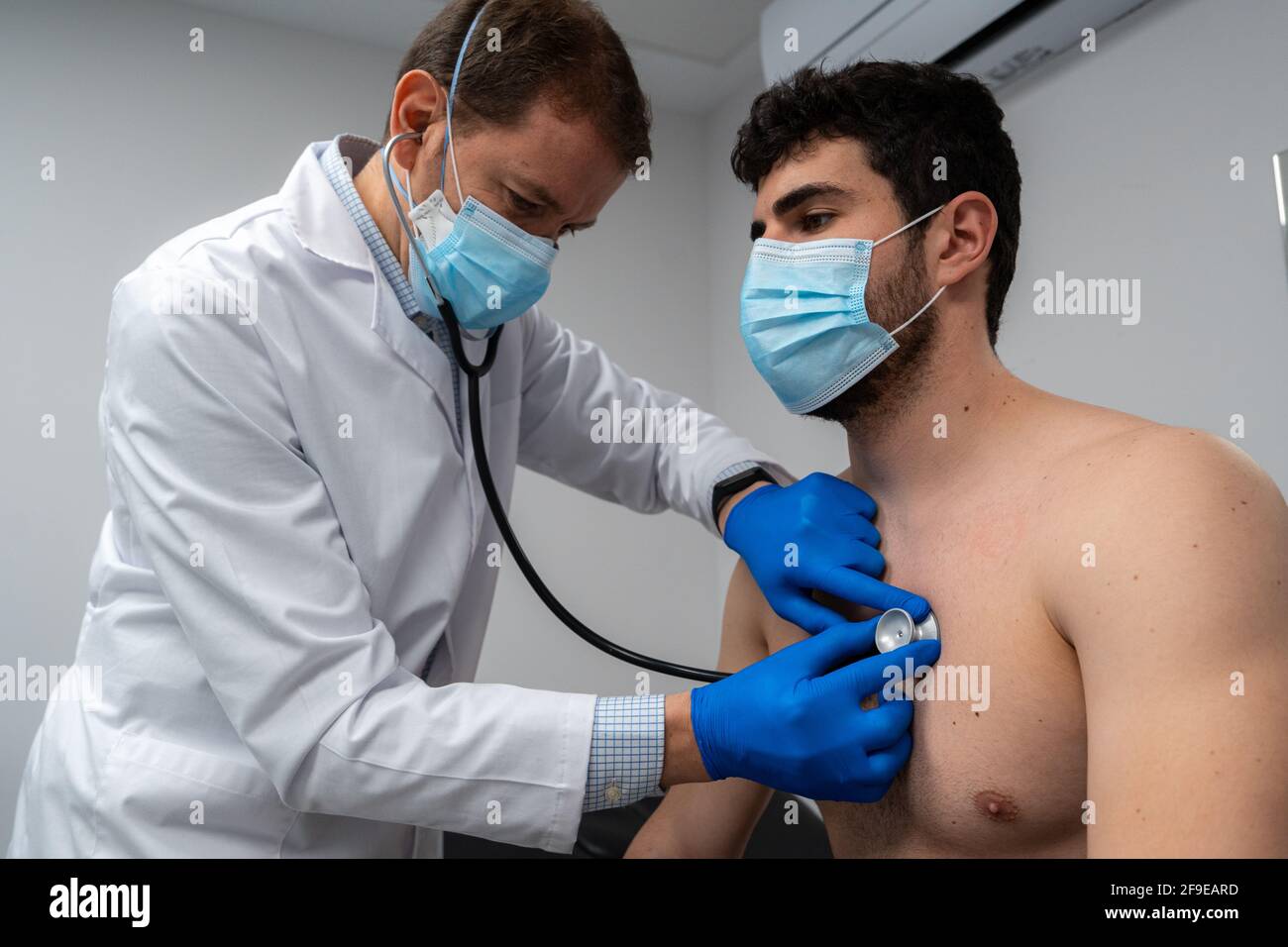 Low angle of male physician in mask using phonendoscope while examining lunges of patient in ...