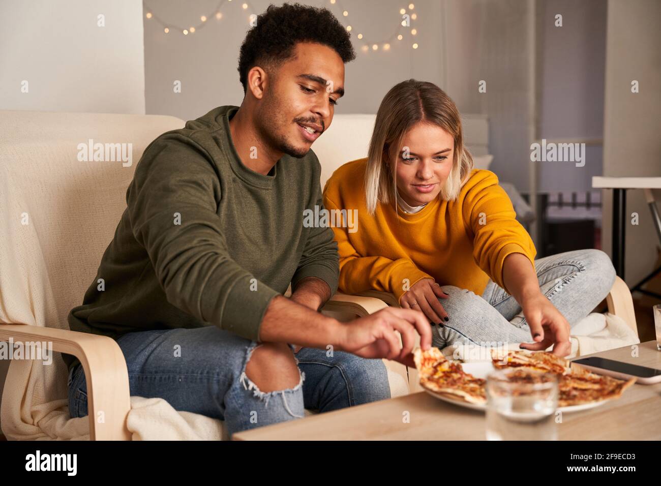 Multiethnic couple sitting in armchairs and eating delicious pizza ...