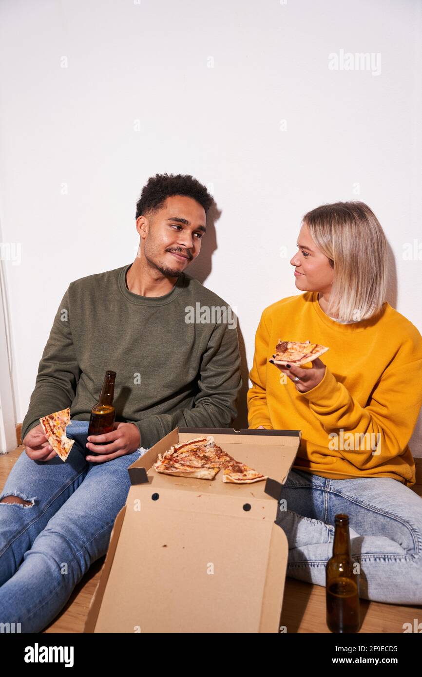 Cheerful multiethnic couple sitting on floor at home while eating ...
