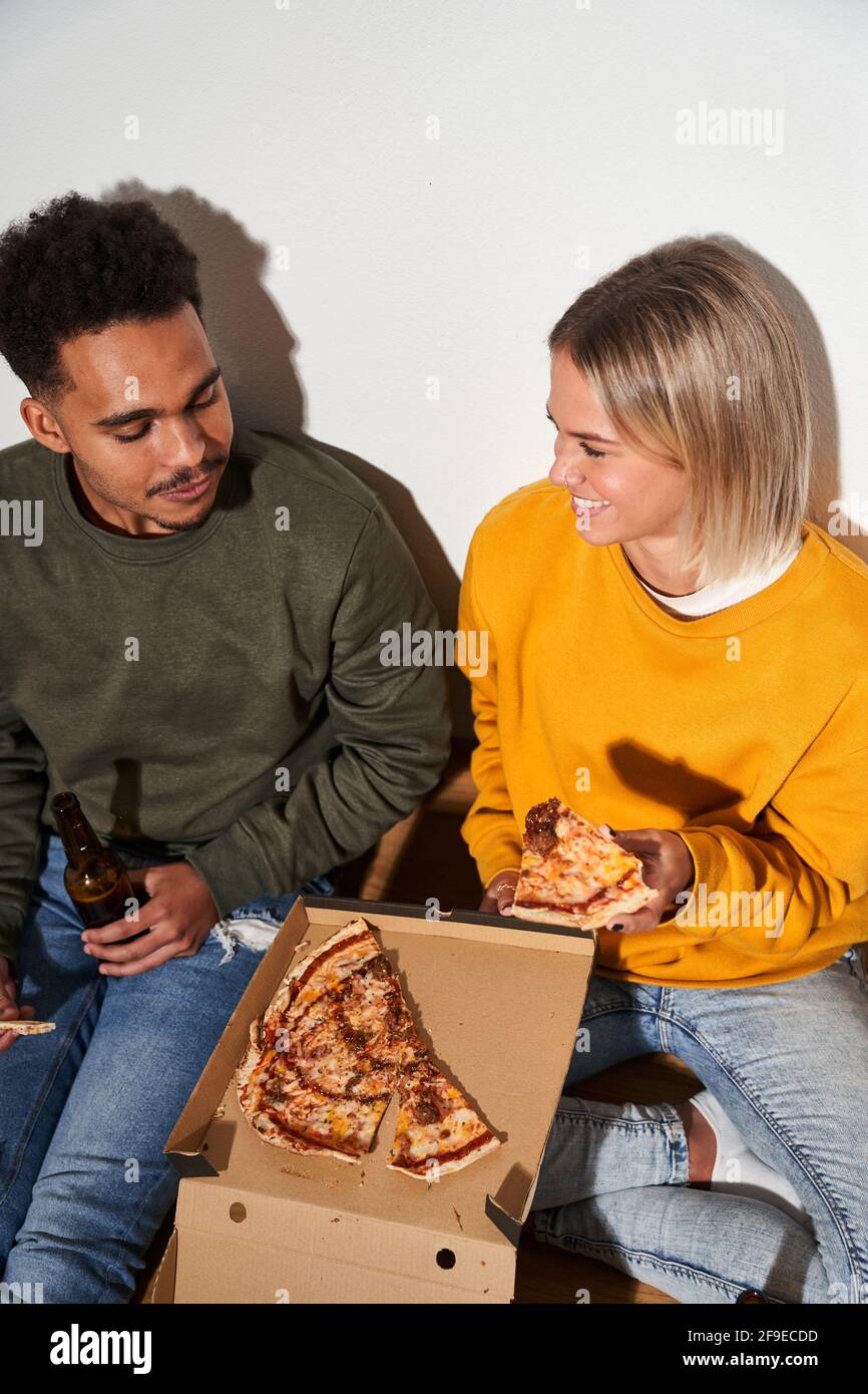 Cheerful multiethnic couple sitting on floor at home while eating ...
